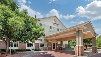 Um hotel Comfort Suites com entrada coberta, fachada de tijolos e uma grande árvore em frente.