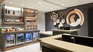 Uma área de recepção moderna com balcão, dispensadores de bebidas geladas, prateleiras com lanches e arte decorativa na parede.