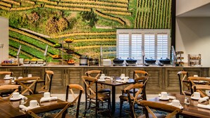 Espaço de café da manhã com um grande mural de um vinhedo, mobiliário de madeira e um buffet com diversos alimentos.