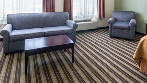 Quarto de hotel com sofá, poltrona e uma pequena mesa. Há uma janela com cortinas e um aparelho de ar condicionado na parede.