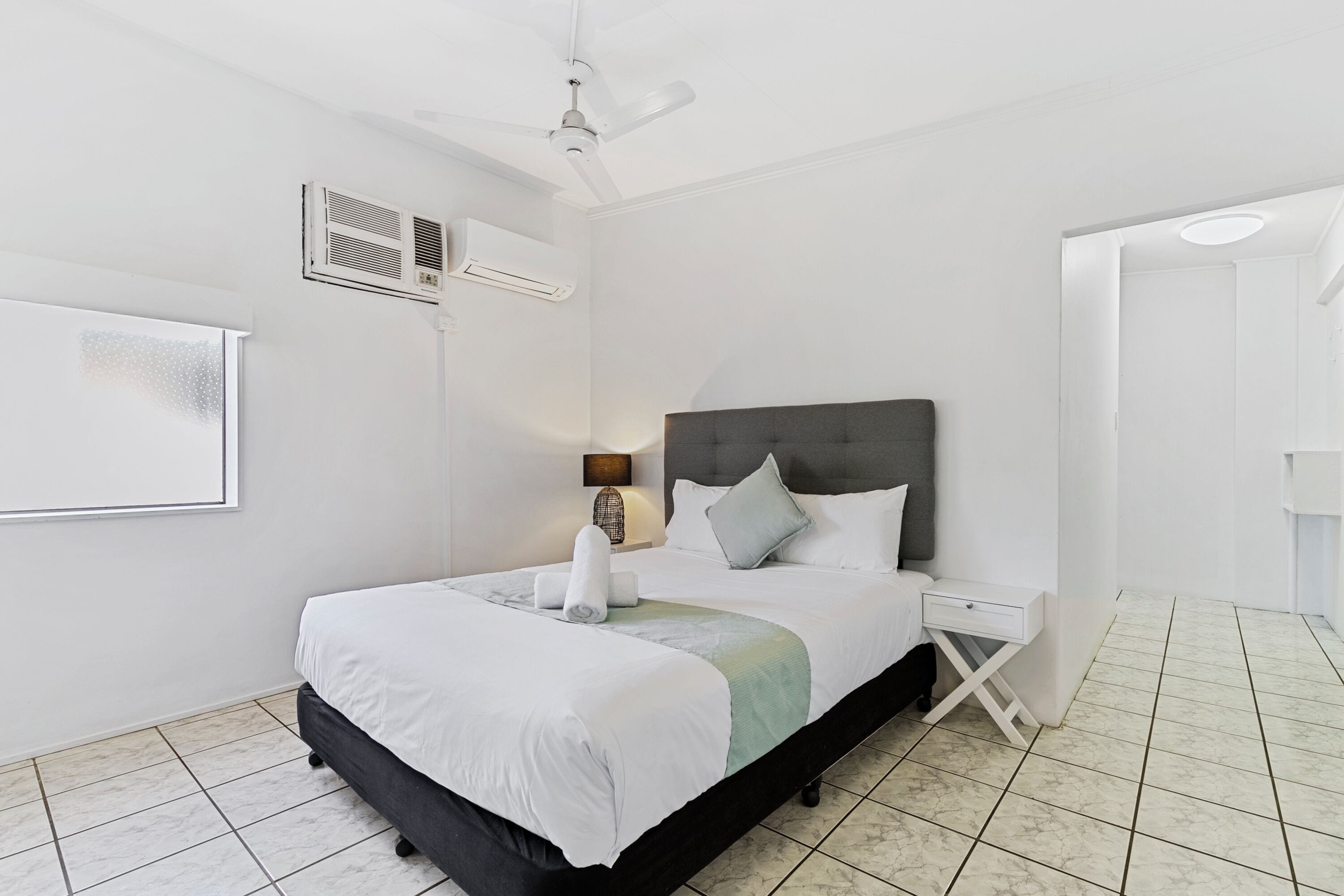 Quarto de hotel moderno com uma cama grande, uma mesa de cabeceira, uma luminária, ar condicionado e um ventilador de teto.