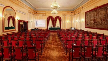 Uma sala de conferências com fileiras de cadeiras vermelhas, piso de madeira, um candelabro e uma grande tapeçaria na parede.