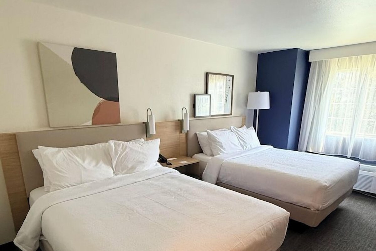 Quarto de hotel com duas camas, uma mesa de cabeceira, uma luminária e um grande quadro abstrato na parede.