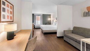 Quarto de hotel com cama, sofá, escrivaninha com luminária, cadeira e um quadro na parede.