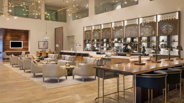 Um hall de hotel espaçoso e moderno, com um balcão de recepção extenso, área de estar confortável e bar com prateleiras exibindo diversas bebidas.