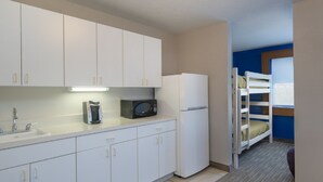 Cozinha compacta com armários brancos, micro-ondas e geladeira. No quarto ao lado, há uma cama beliche com janela.