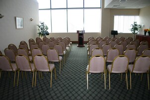 Uma sala de conferências com fileiras de cadeiras voltadas para um púlpito, grandes janelas e piso coberto de carpete.