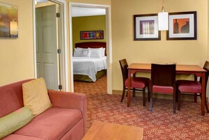 Quarto de hotel com sofá vermelho, mesa de jantar de madeira com quatro cadeiras, cama com roupa de cama branca e um quadro colorido na parede.