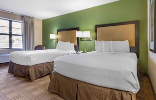 Quarto de hotel com duas camas, uma janela com cortinas, uma cadeira e uma mesa com luminária.