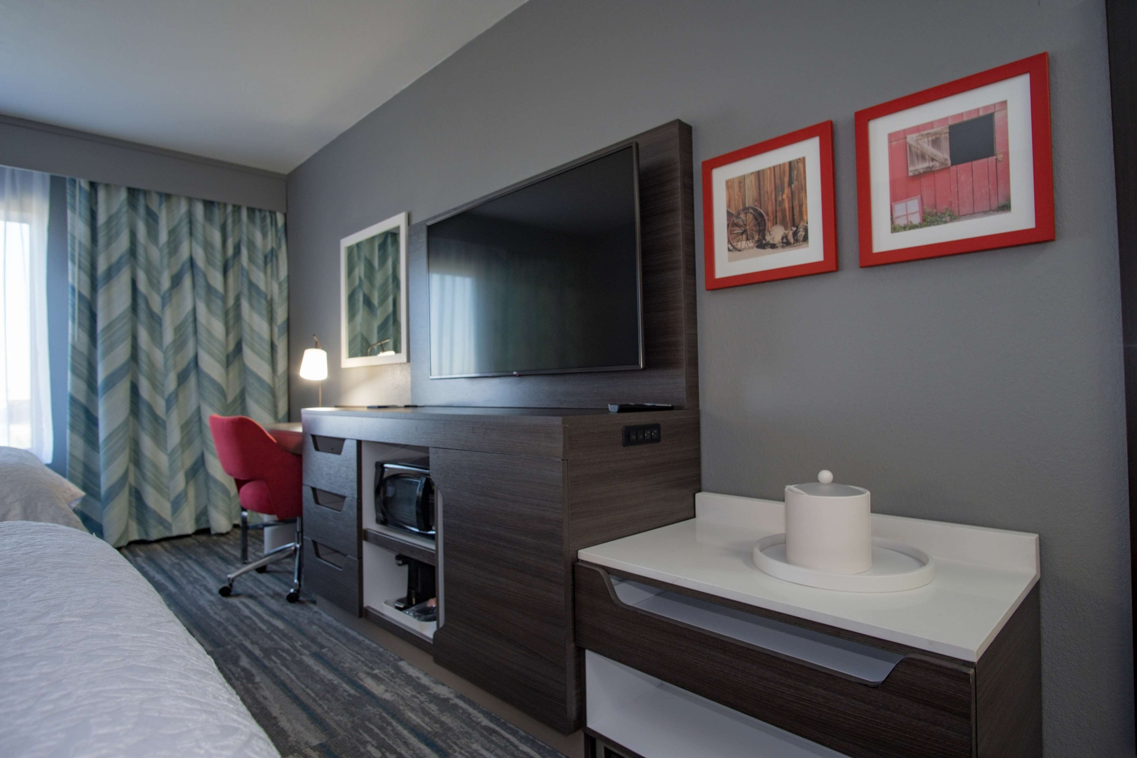 Quarto de hotel com cama, escrivaninha com cadeira, televisão de tela plana, prateleira pequena e duas fotografias emolduradas na parede.