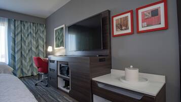 Quarto de hotel com cama, escrivaninha com cadeira, televisão de tela plana, prateleira pequena e duas fotografias emolduradas na parede.