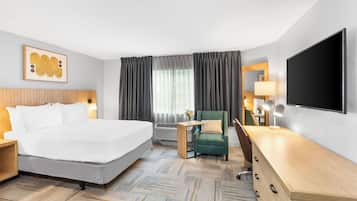 Quarto de hotel com uma cama grande, escrivaninha, cadeira, televisão e janela com cortinas.