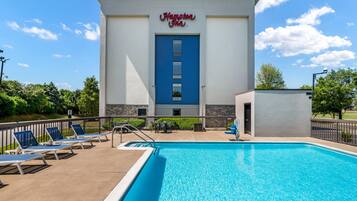 Piscina com cadeiras de descanso, placa da Hampton Inn e céu azul claro.