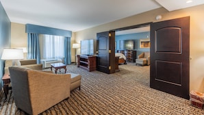Quarto de hotel com sofá, poltrona, televisão e escrivaninha com luminária.