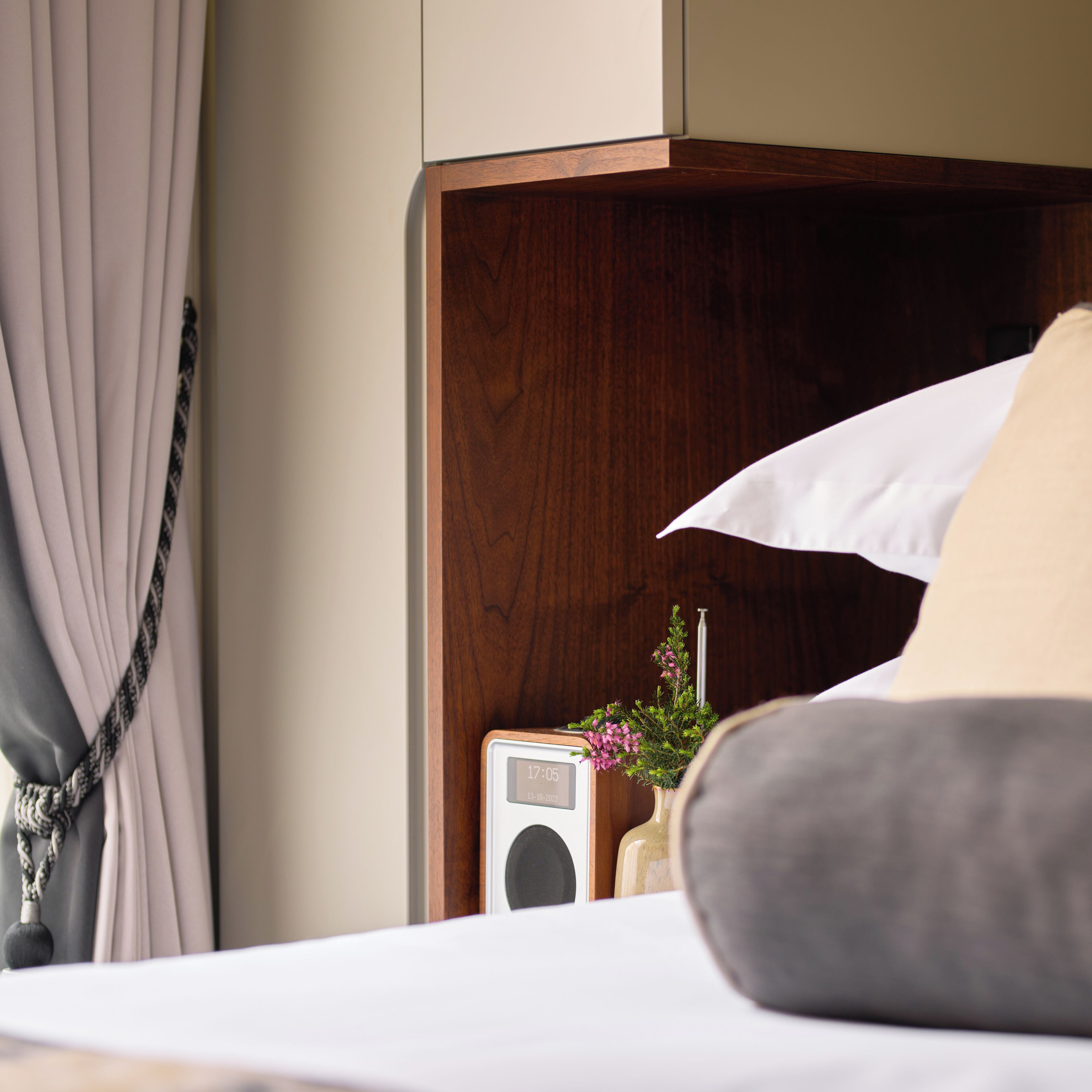 Cama bem arrumada com travesseiro branco, mesa de cabeceira com rádio e um vaso de flores, e cortina parcialmente aberta.