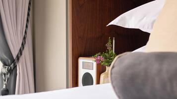 Cama bem arrumada com travesseiro branco, mesa de cabeceira com rádio e um vaso de flores, e cortina parcialmente aberta.