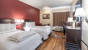 Quarto de hotel com duas camas, uma escrivaninha, uma cadeira, uma luminária de mesa, um quadro na parede e uma janela com cortinas.