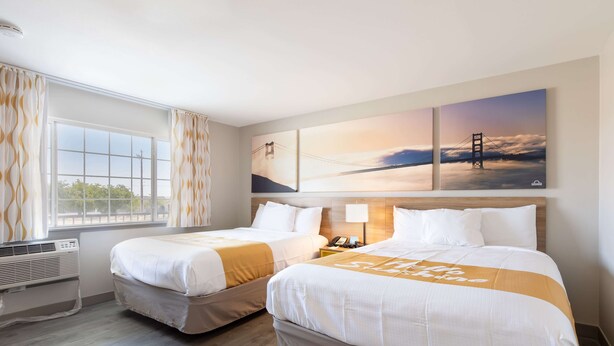 Quarto de hotel com duas camas, uma janela com cortinas, uma obra de arte pendurada na parede retratando uma ponte e uma mesa de cabeceira com abajur.