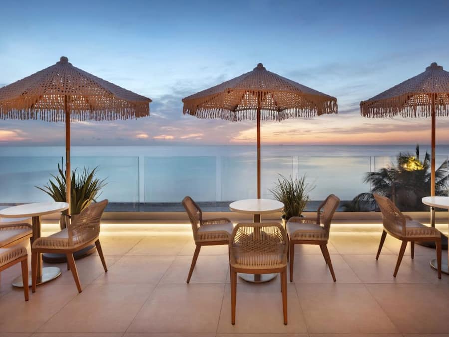 Terraço com móveis de vime e guarda-sóis de palha, com vista para um mar calmo.