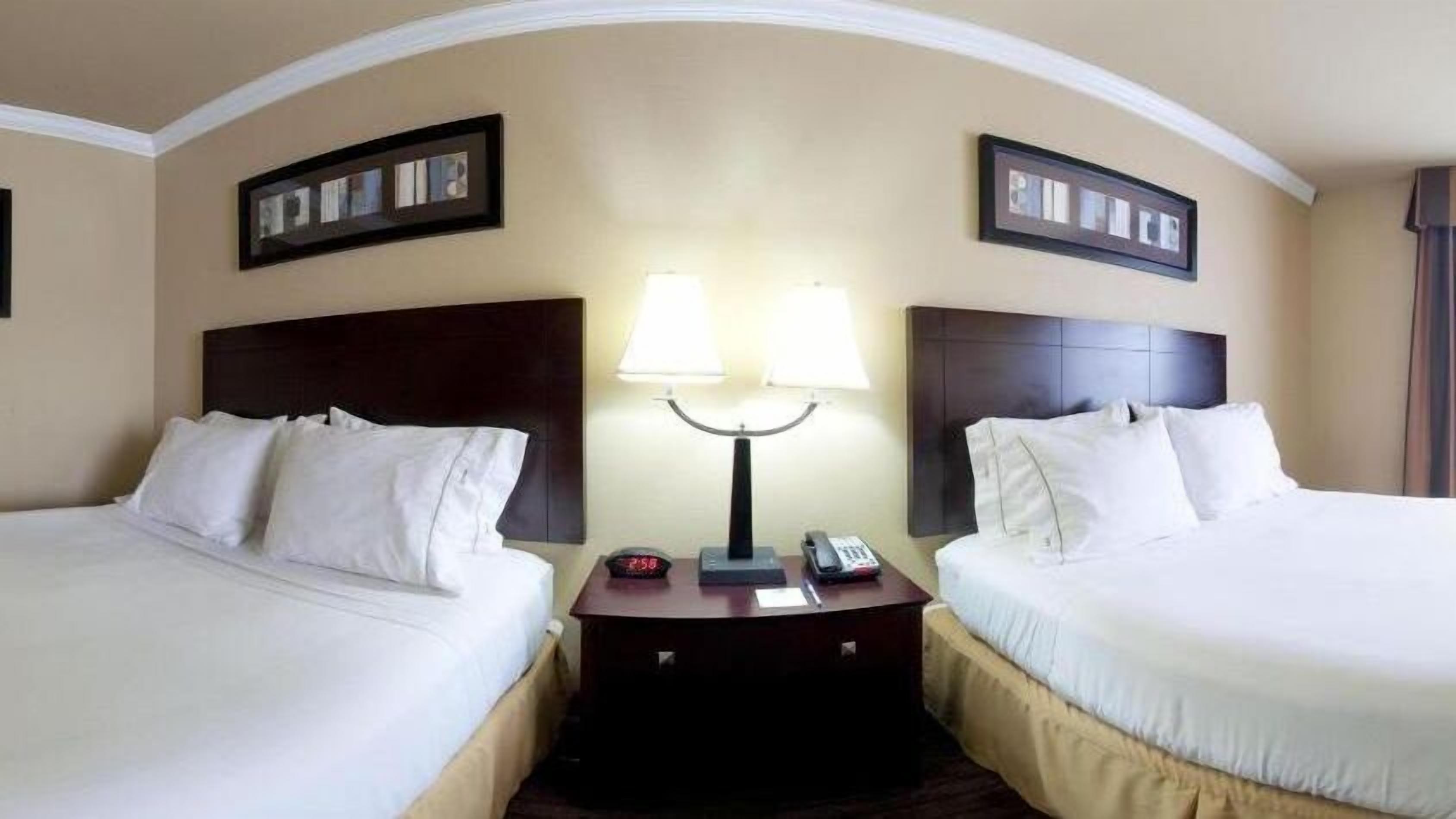 Quarto de hotel com duas camas, mesa de cabeceira com luminária e quadros nas paredes.
