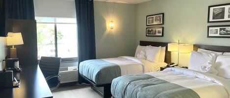 Quarto de hotel com duas camas, uma escrivaninha, uma cadeira, um abajur e uma janela com cortinas.