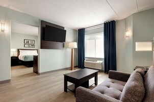 Quarto de hotel com televisão de tela plana na parede, cama com cabeceira, mesa de cabeceira com abajur, área de estar com sofá e poltrona, e uma janela grande com cortinas.