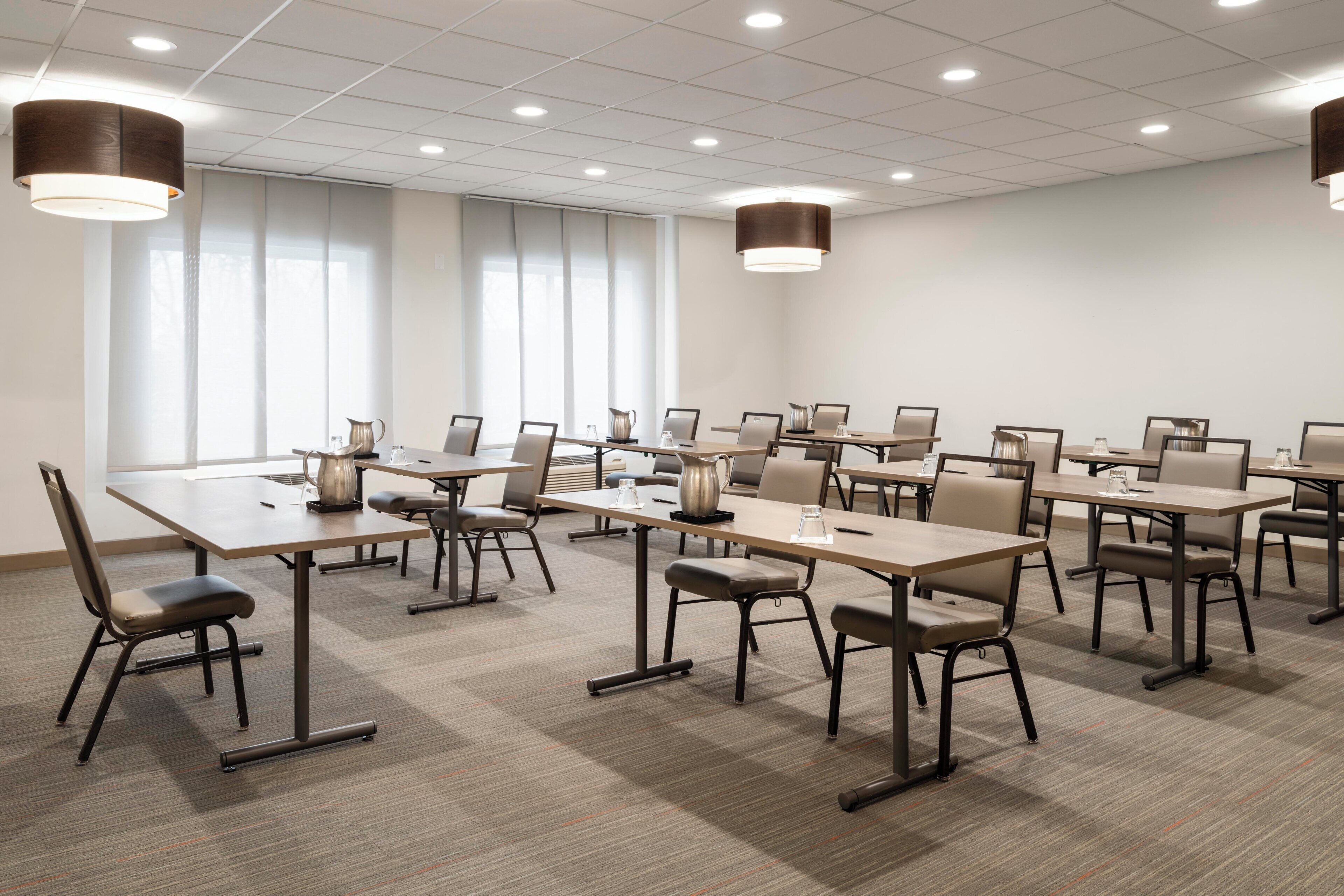 Uma sala de conferências com várias mesas e cadeiras, janelas amplas com persianas e luminárias no teto.