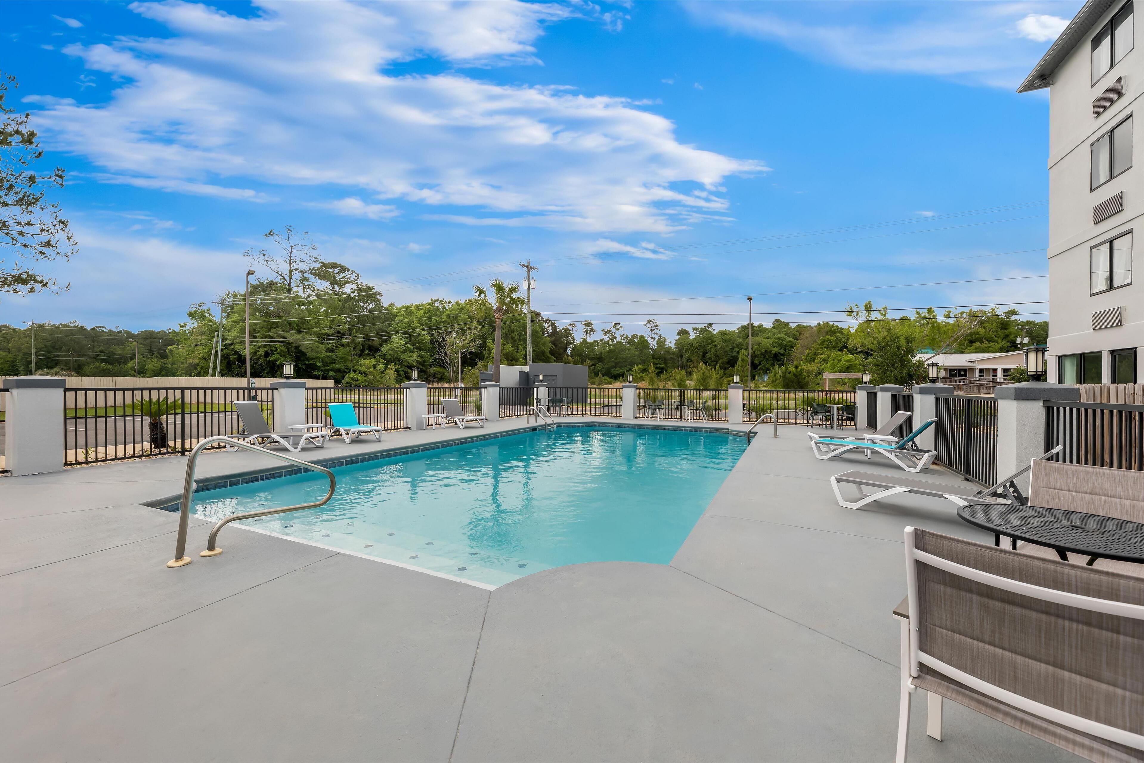 Piscina com espreguiçadeiras e área de estar coberta, cercada por uma vedação e árvores.