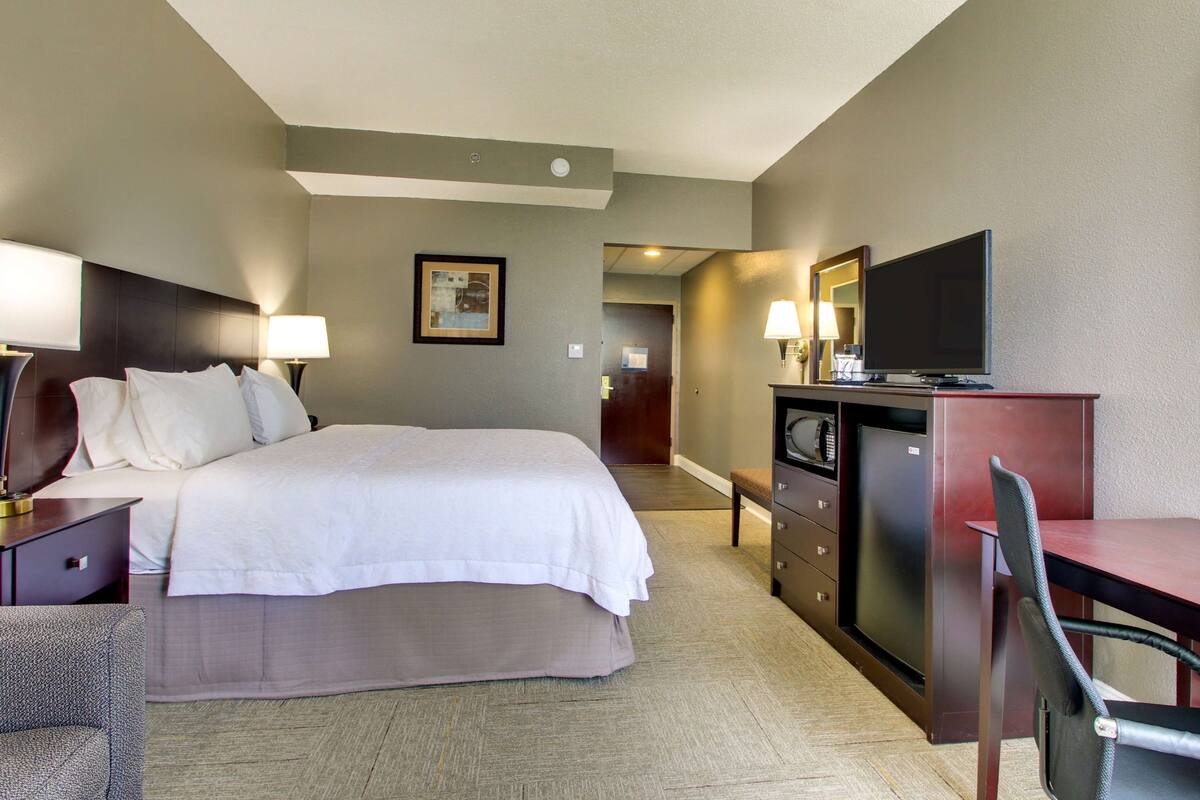 Quarto de hotel com uma cama, abajures, televisão, escrivaninha e uma cadeira.