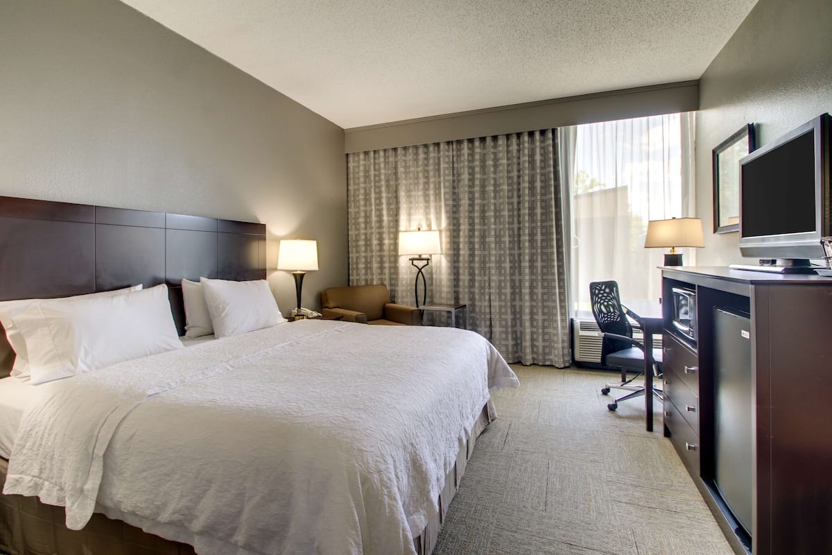 Quarto de hotel com uma cama grande, duas luminárias de cabeceira, uma escrivaninha com cadeira, uma televisão e uma janela ampla com cortinas.