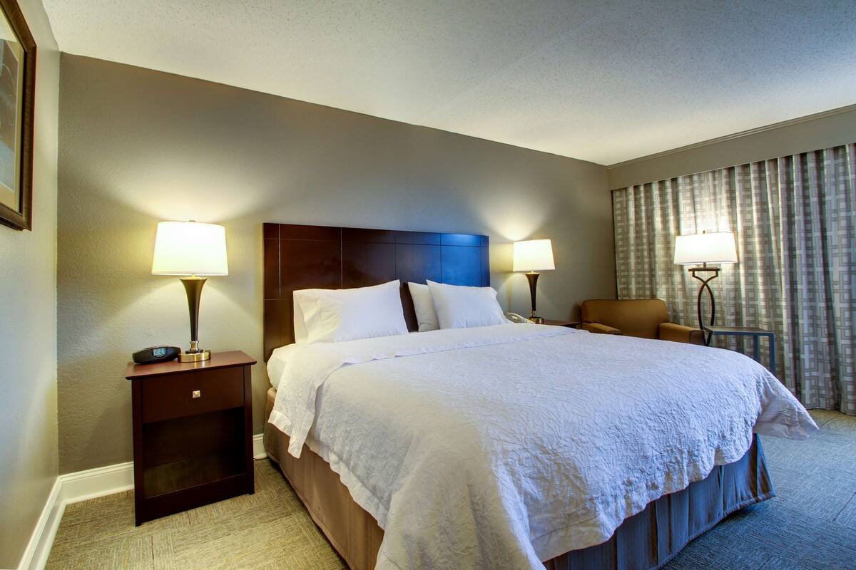Quarto de hotel com uma cama grande, duas mesas de cabeceira com luminárias, uma cadeira e uma cortina.