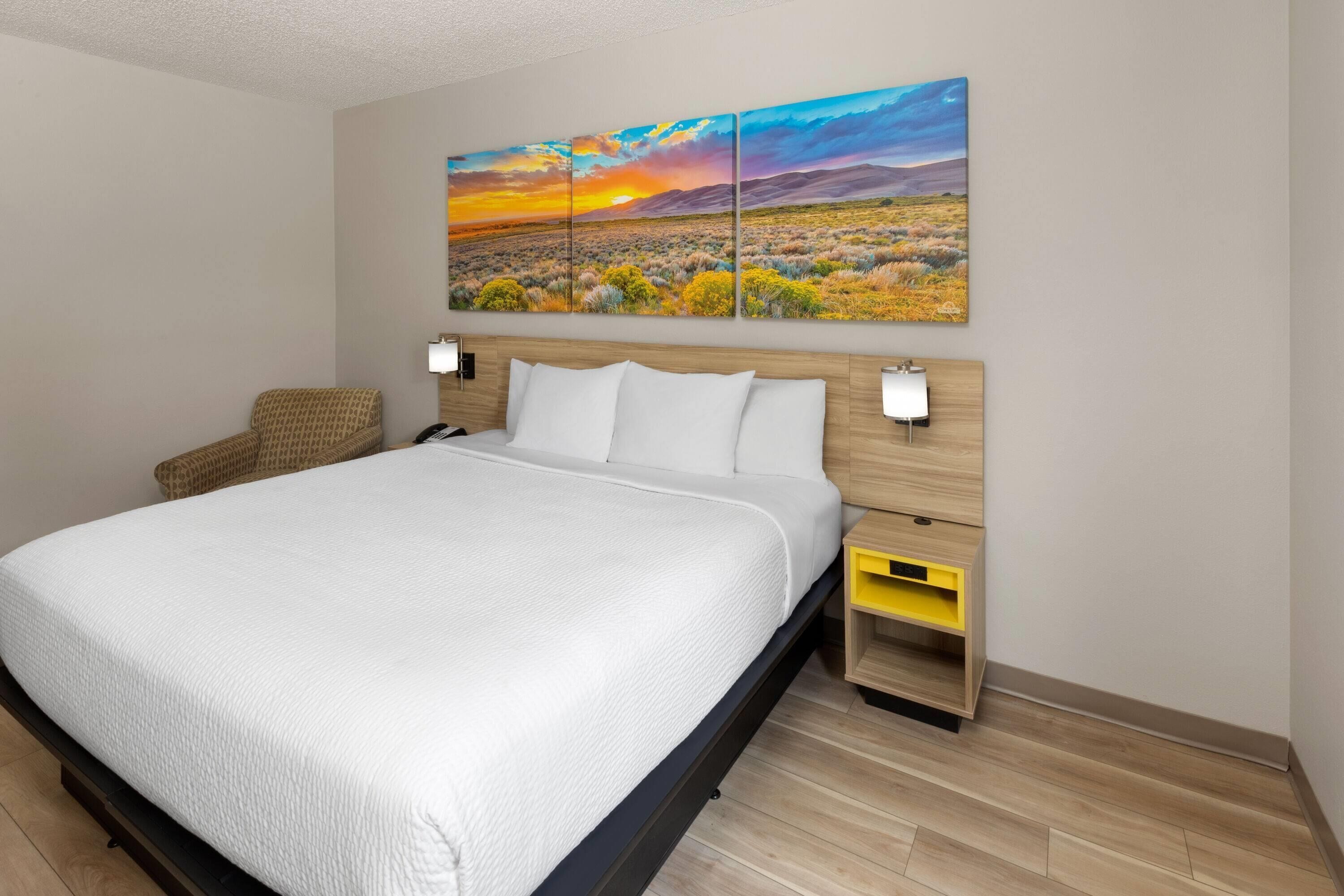 Quarto com uma cama grande, cabeceira de madeira, duas mesas de cabeceira com luminárias e um quadro na parede com paisagem desértica.