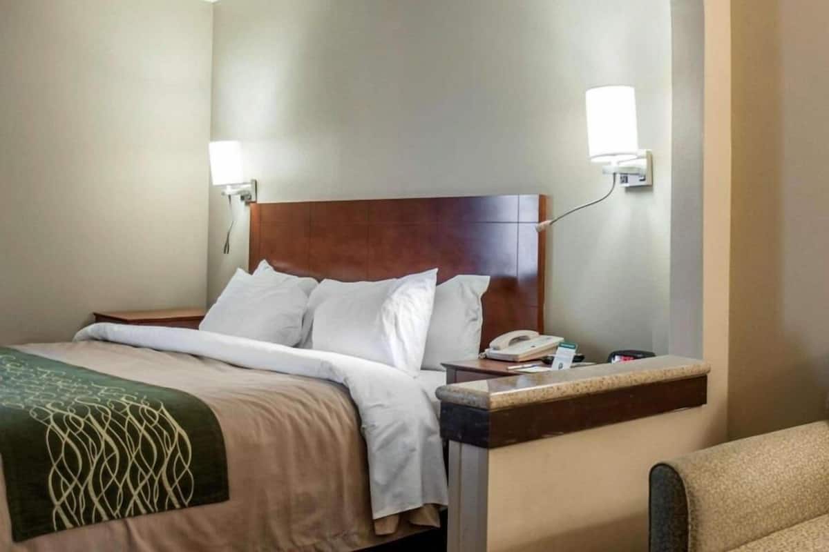 Quarto de hotel com cama, mesa de cabeceira, luminária e uma cadeira.