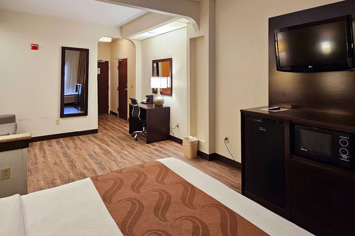 Quarto de hotel com cama, escrivaninha com cadeira, televisão, micro-ondas e uma pequena lixeira.