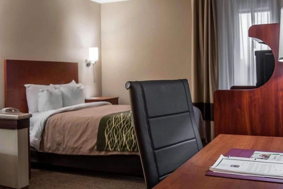 Quarto de hotel com uma cama, uma escrivaninha com cadeira, uma luminária e uma janela com cortinas.