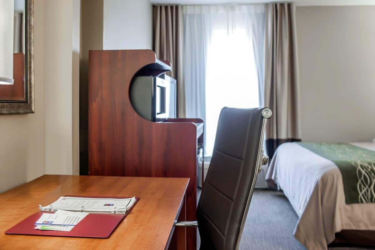 Quarto de hotel com escrivaninha de madeira, uma cadeira, cama com colcha verde e janela com cortinas.