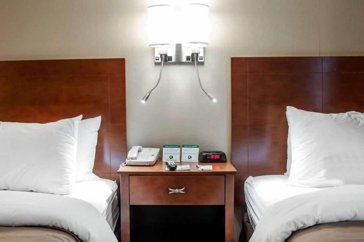 Quarto de hotel com duas camas, uma mesa de cabeceira com telefone, relógio e gavetinha.