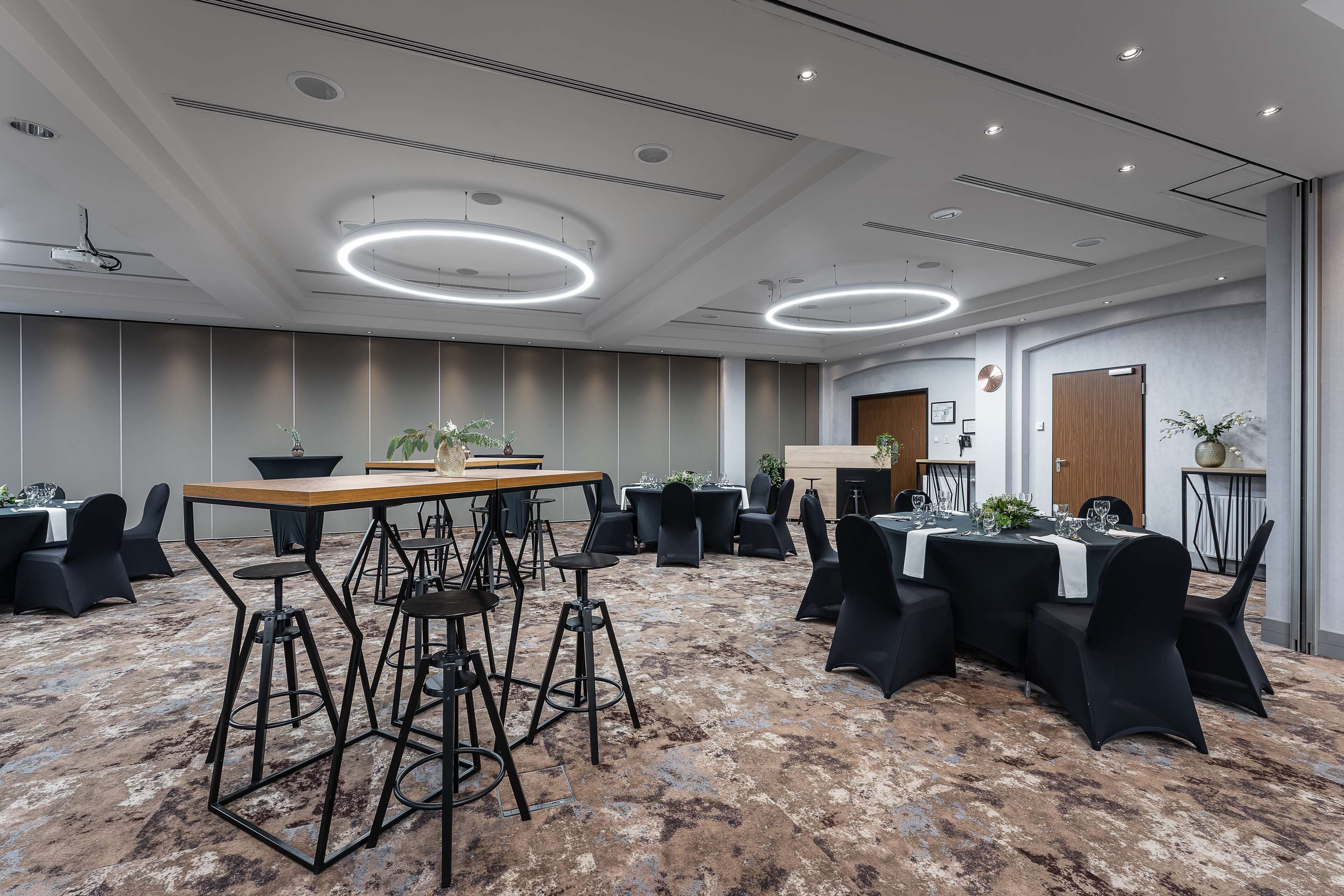 Sala de conferências com mesas redondas, cadeiras pretas e piso coberto por carpete.