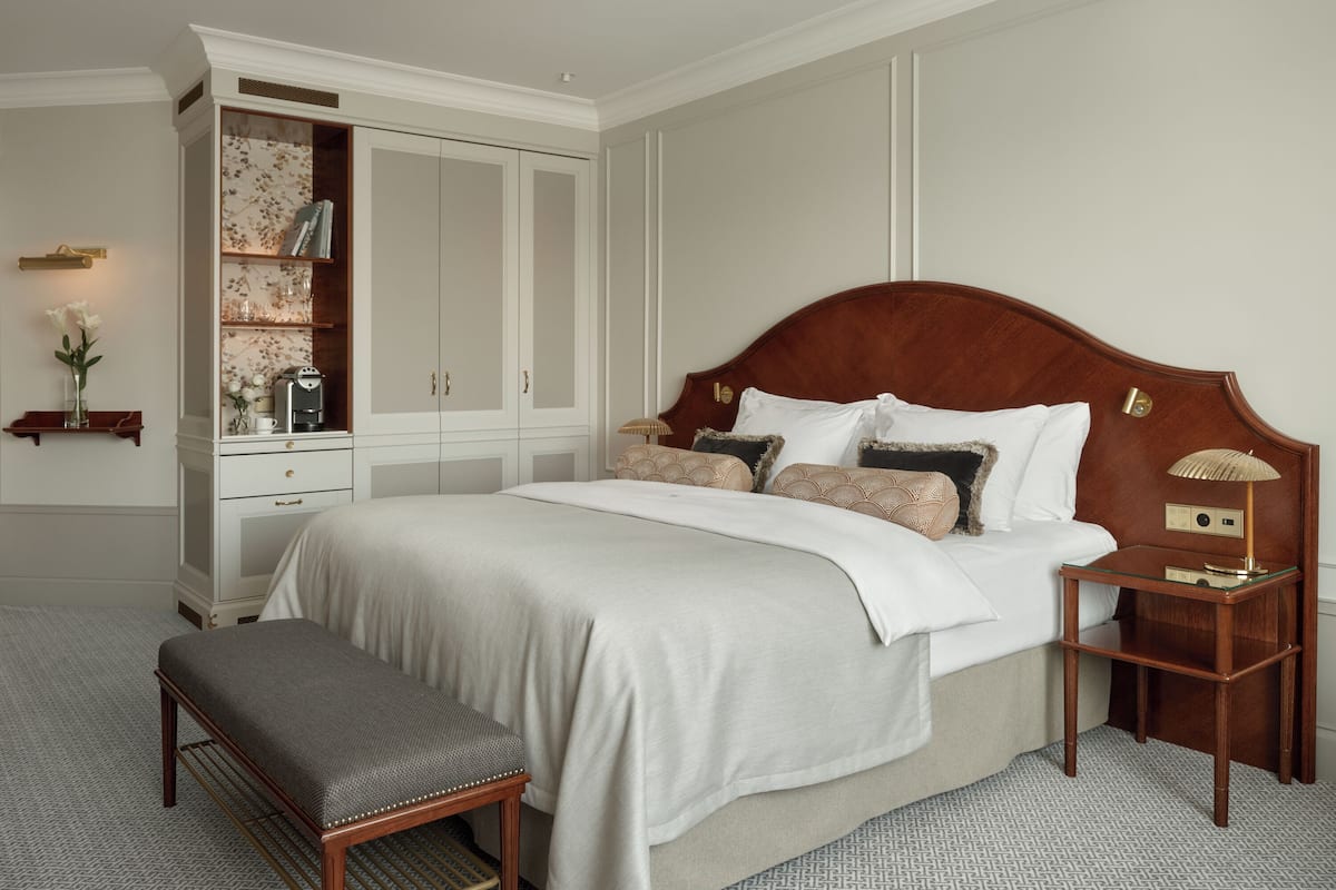 Quarto com cama grande, mesa de cabeceira de madeira, um banco, guarda-roupa e prateleira com objetos decorativos.