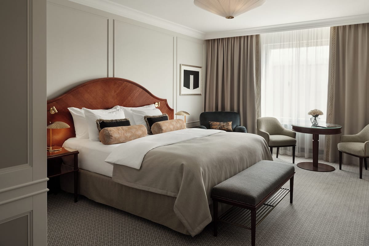 Quarto de hotel com uma cama grande, um banco, duas cadeiras, uma mesa pequena e um lustre.