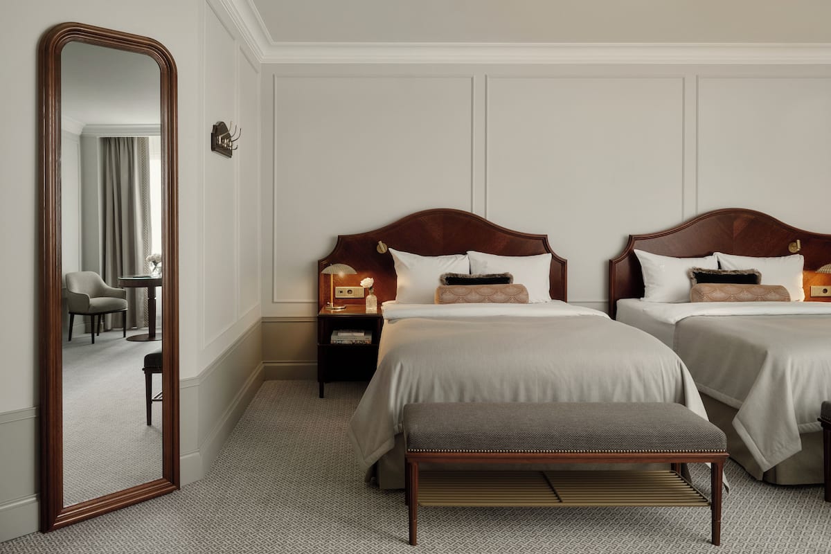 Quarto de hotel com duas camas, um espelho grande, um banco, uma mesa de cabeceira com abajur e vista para um corredor com cadeira e cortinas.