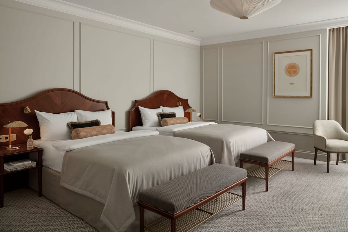 Quarto de hotel com duas camas, um banco, uma cadeira, uma mesinha de cabeceira com abajur e um quadro na parede.