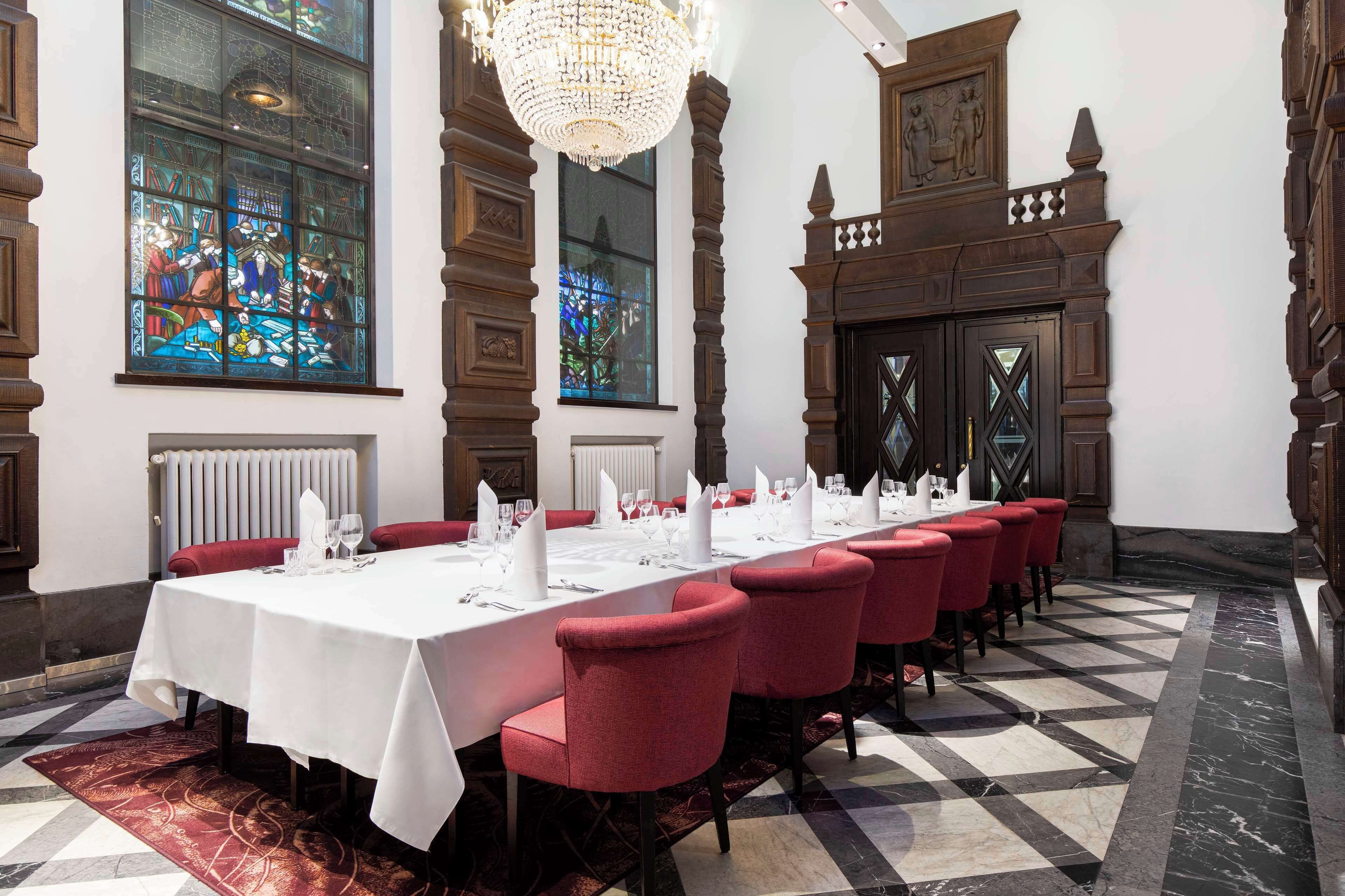 Uma sala de jantar com uma mesa comprida posta para refeição, cadeiras estofadas em vermelho, um lustre, janelas de vitrais e uma lareira.