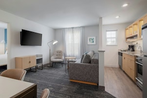 Quarto de hotel moderno com sofá, poltrona, mesa de jantar, televisão de tela plana, cozinha compacta e uma grande janela com cortinas.