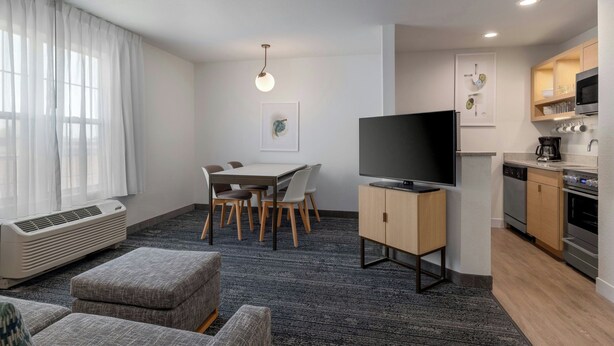 Uma sala moderna com televisão de tela plana, área de jantar com mesa e cadeiras, cozinha compacta com micro-ondas e forno, e uma janela grande com cortinas translúcidas.