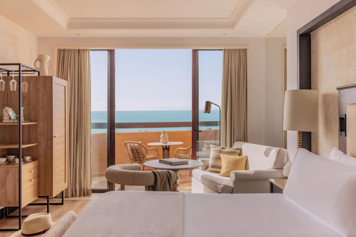 Quarto de hotel com cama, sofá, mesa pequena e cadeira. Grande janela com vista para o mar.