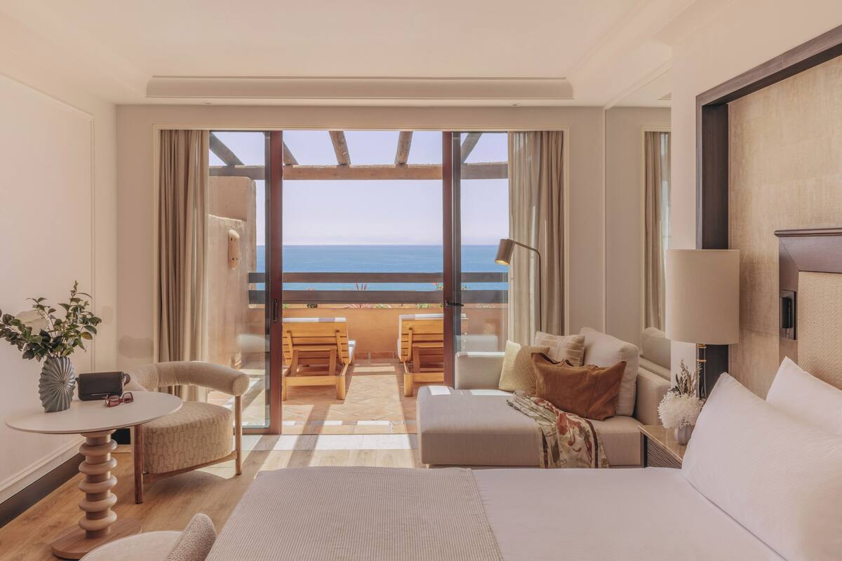 Quarto de hotel com cama, sofá, poltrona, mesa pequena e vista para o oceano.
