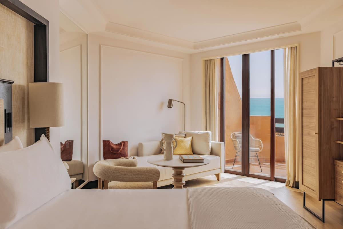Quarto de hotel com cama, mesa redonda, sofá, poltrona, abajur e vista para o mar por grandes janelas.