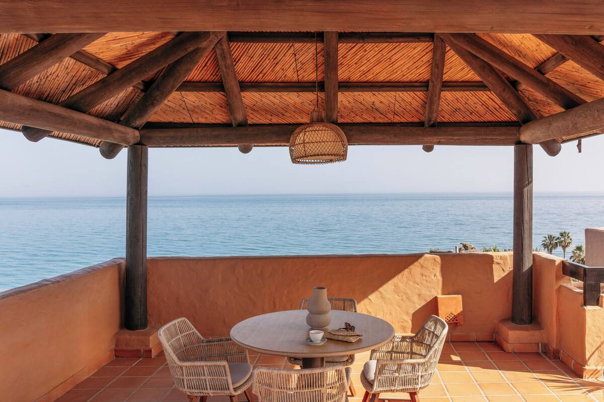 Uma área de estar coberta ao ar livre, com mesa, cadeiras e um lustre pendurado, com vista para o mar.