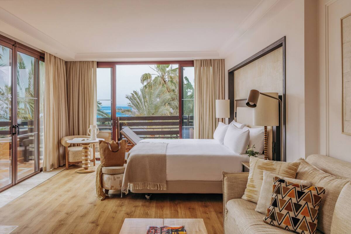 Quarto de hotel com uma cama grande, um sofá, uma mesa pequena, uma luminária e vista para o mar.